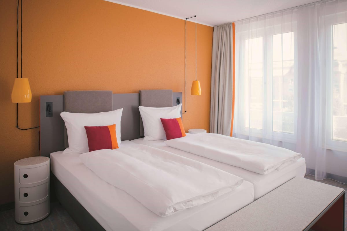 Uma cama de casal com roupa de cama branca e duas almofadas, uma vermelha e outra laranja, num quarto com paredes laranja e uma janela com cortinas translúcidas.