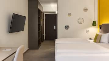 Quarto de hotel com cama, escrivaninha e televisão. Há decorações nas paredes e uma porta que dá para outro cômodo.