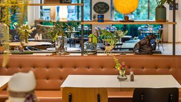 Interior de restaurante com um banco estofado em castanho, mesas de madeira e prateleiras com plantas e objetos decorativos.
