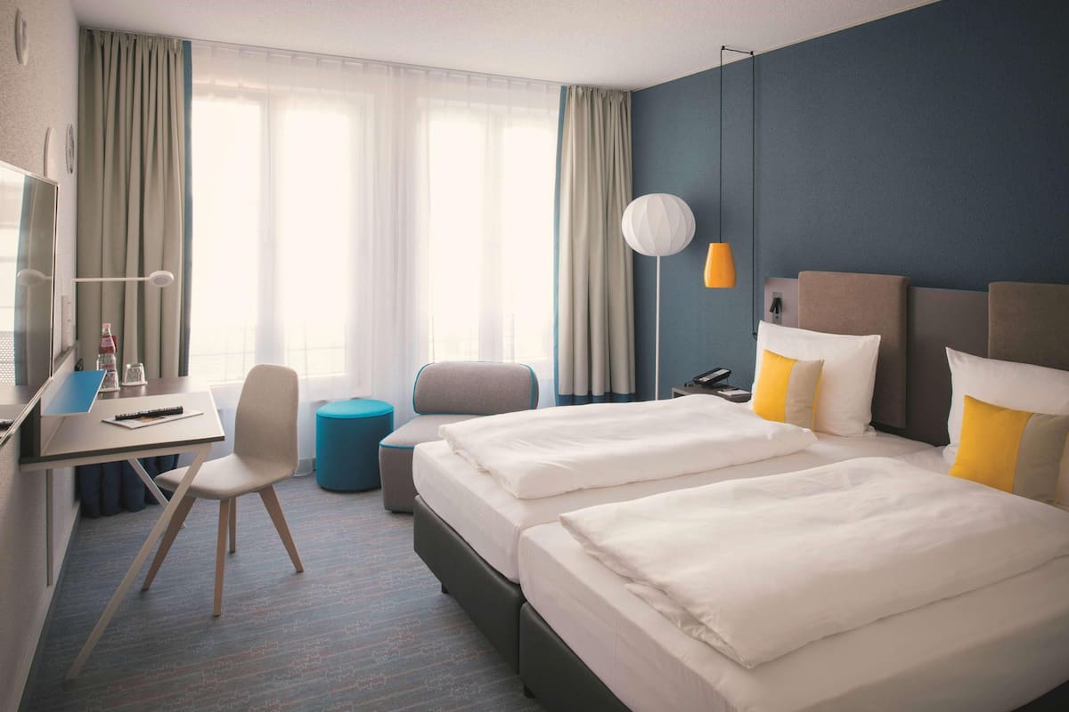 Quarto de hotel com duas camas, uma escrivaninha, uma cadeira e um abajur.