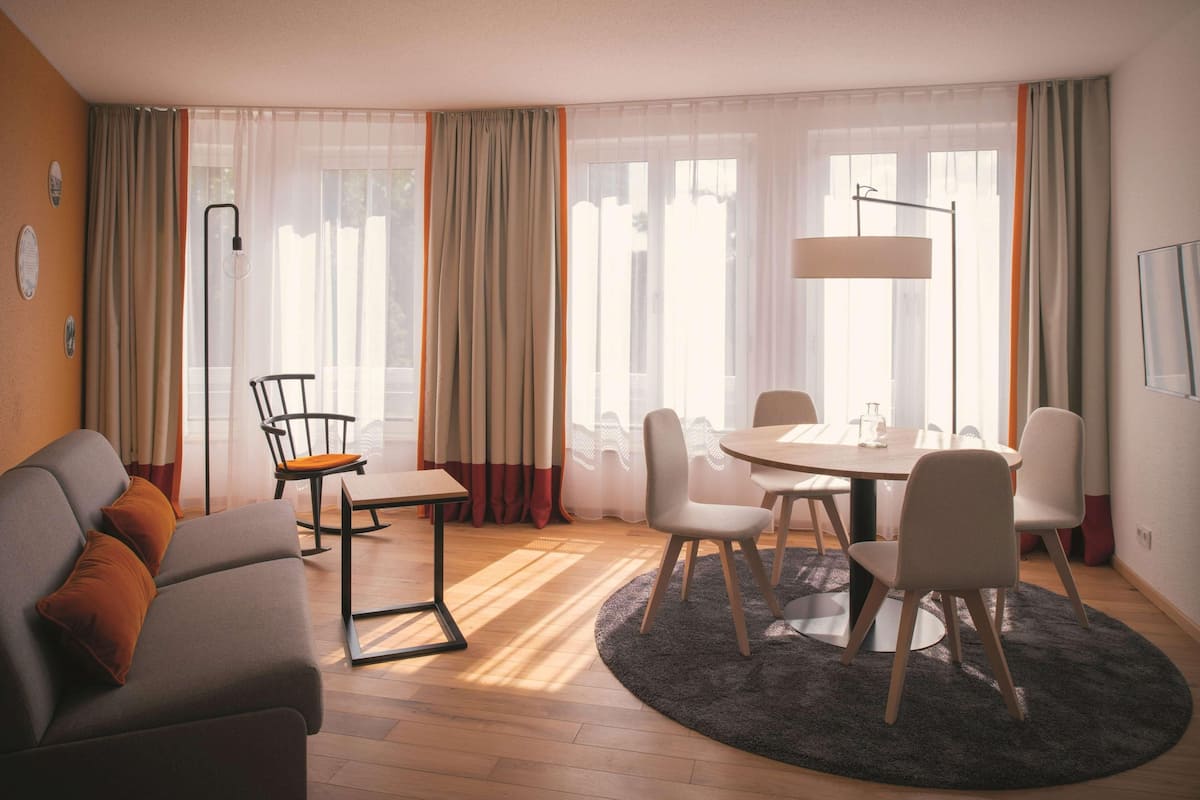 Uma sala de estar moderna com sofá, mesa de jantar com cadeiras, luminária de chão e grandes janelas com cortinas.