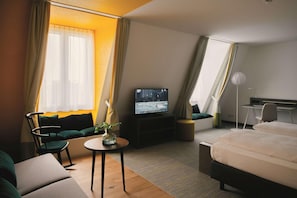Quarto de hotel com cama, televisão, escrivaninha, cadeira, sofá e janela com cortinas.
