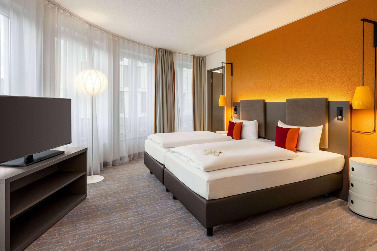 Quarto de hotel com uma cama grande, televisão, mesa de cabeceira e abajur.