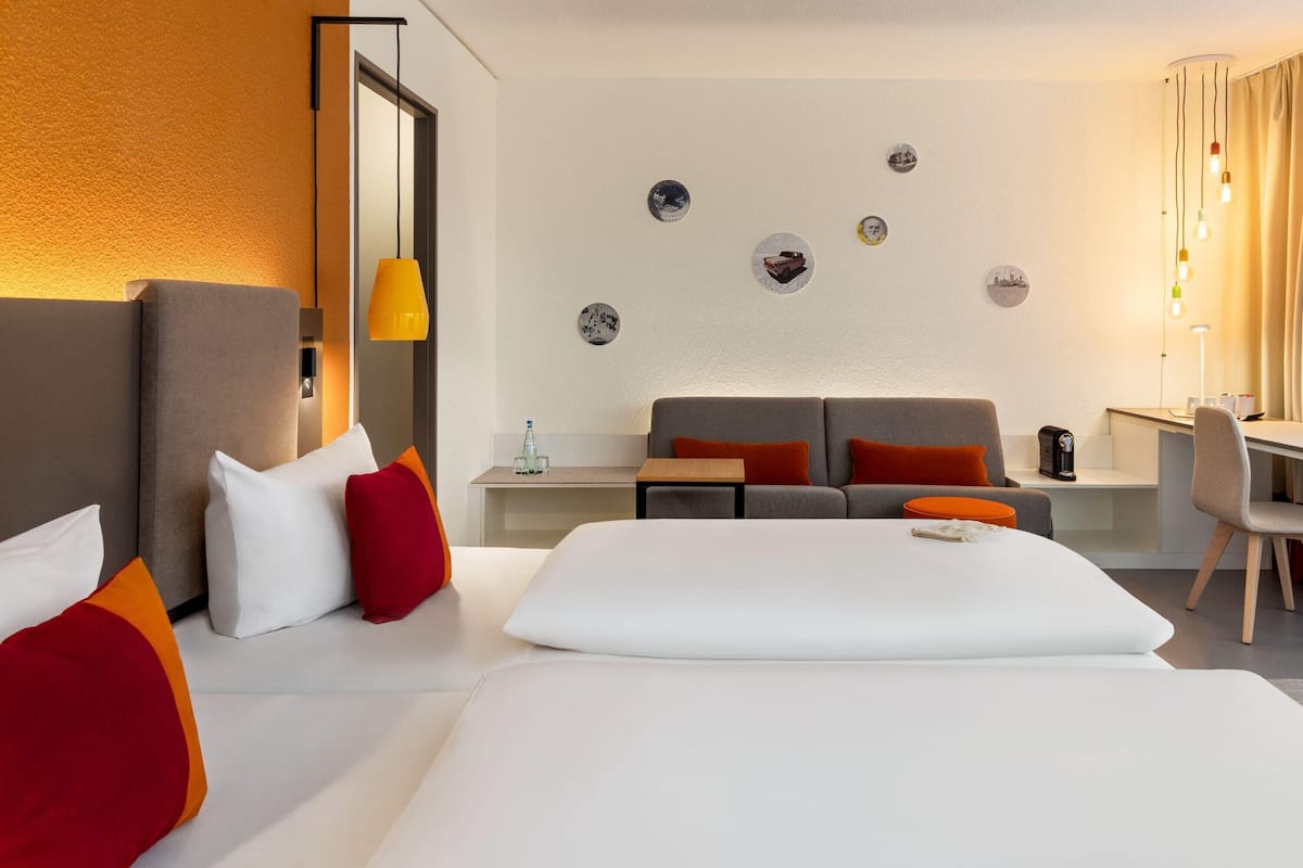 Quarto de hotel moderno com uma cama grande, um sofá, uma escrivaninha e uma cadeira. Há decorações nas paredes e um abajur.