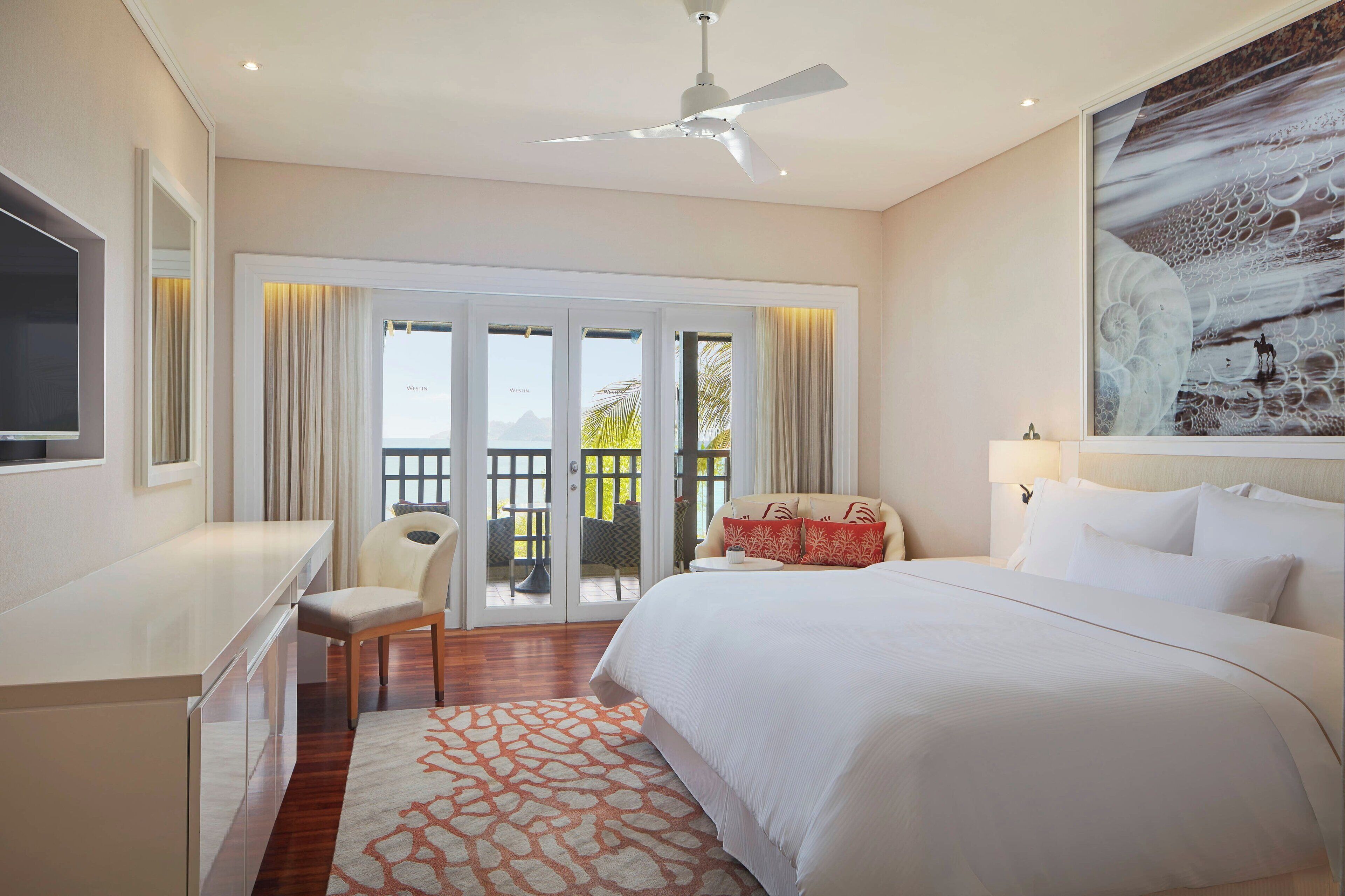 Quarto de hotel com cama, escrivaninha, cadeira, ventilador de teto e vista para o exterior.