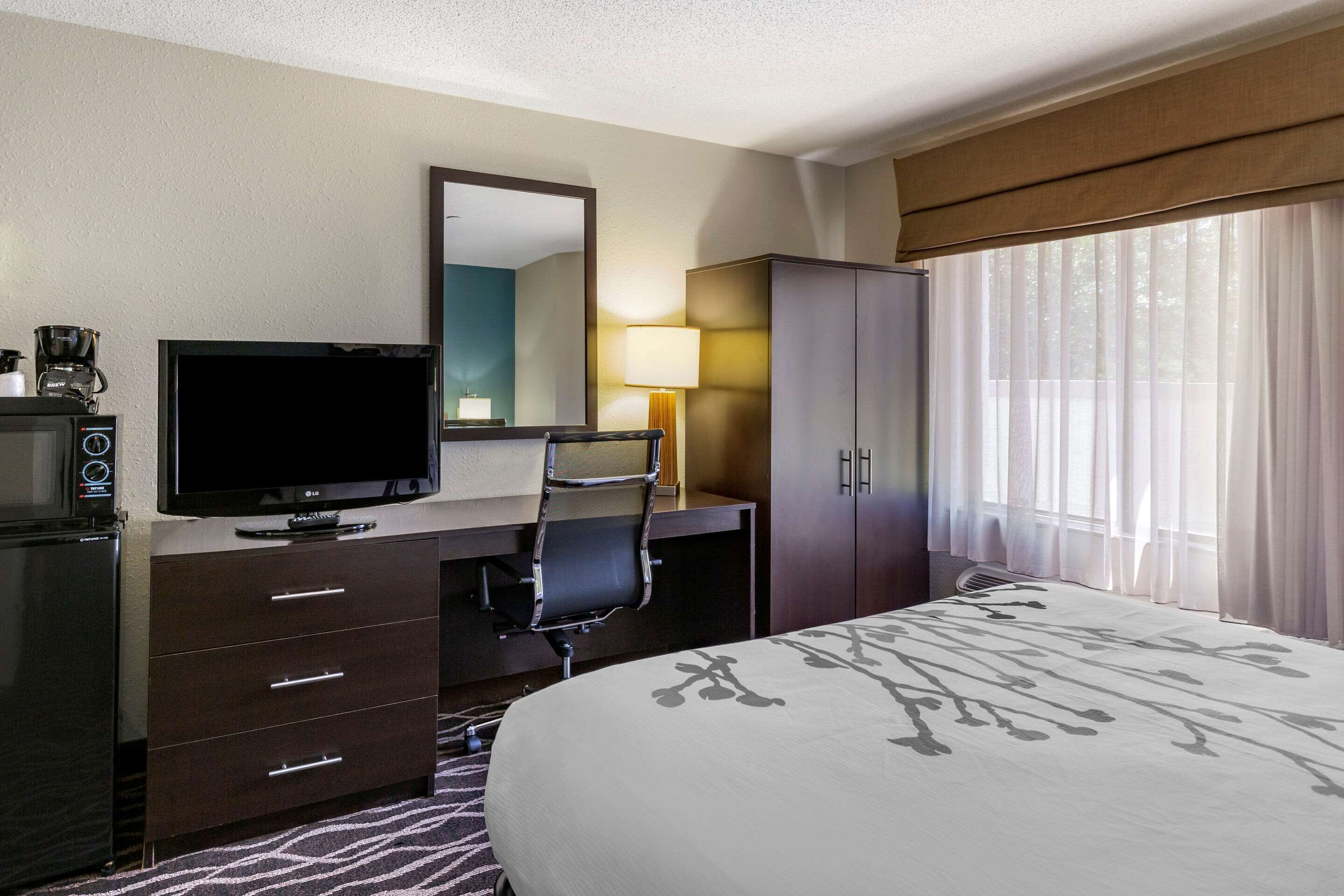 Quarto de hotel com cama, escrivaninha com televisão, cadeira, armário e janela com cortinas.