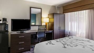 Quarto de hotel com cama, escrivaninha com televisão, cadeira, armário e janela com cortinas.