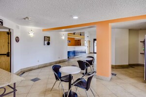 Um saguão de hotel com uma mesa de recepção, uma área de refeições com mesas e cadeiras e uma cozinha ao fundo.