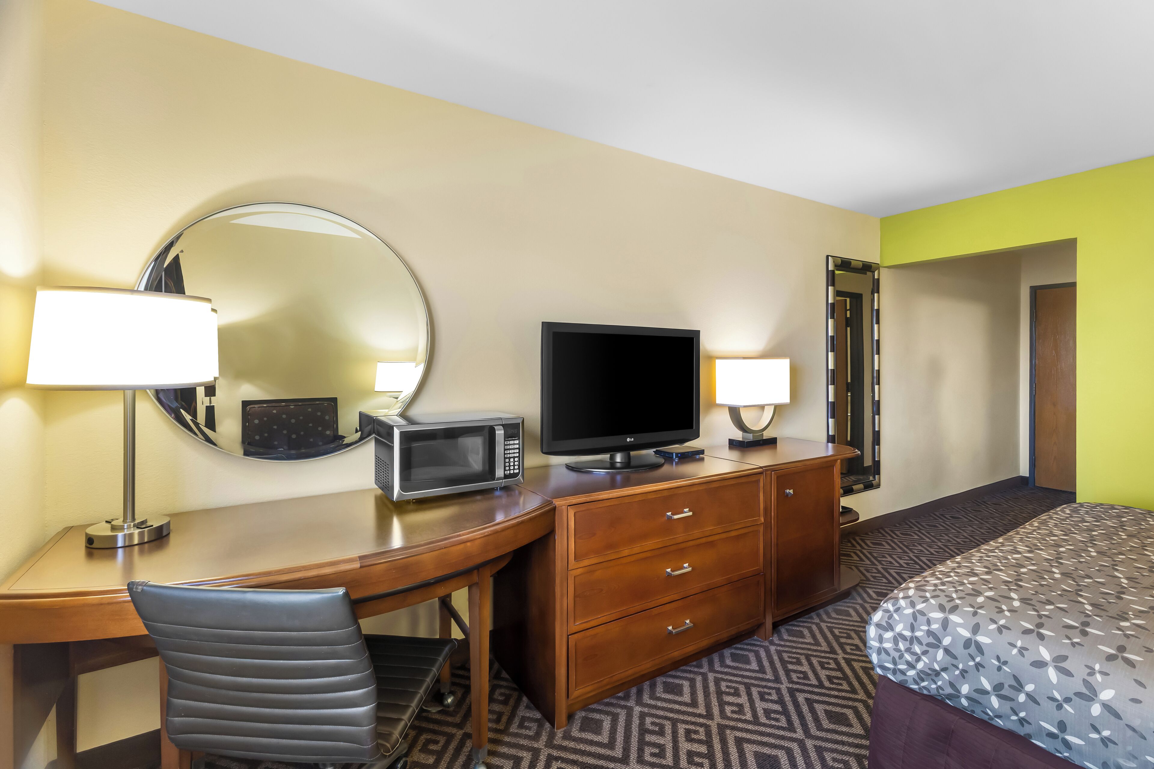 Quarto de hotel com escrivaninha de madeira, televisão, espelho, cama com colcha estampada e tapete estampado.