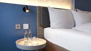 Quarto de hotel moderno com parede azul, mesa de cabeceira redonda com luminária, cama com roupa de cama branca e tomada na parede.