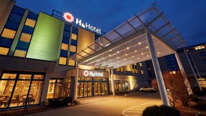 Um hotel moderno com cobertura de entrada em vidro, um destaque para a placa 'H4 Hotel' e fachada bem iluminada.