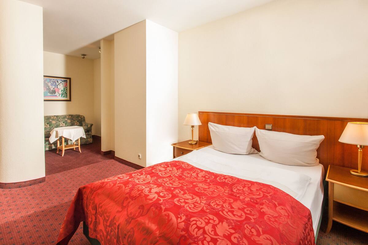Quarto de hotel com uma cama grande, duas mesas de cabeceira com abajures, uma pequena mesa com toalha branca e um sofá floral.