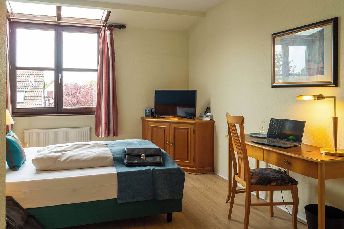 Quarto de hotel com uma cama, uma escrivaninha com um laptop, uma televisão e uma janela com cortinas.