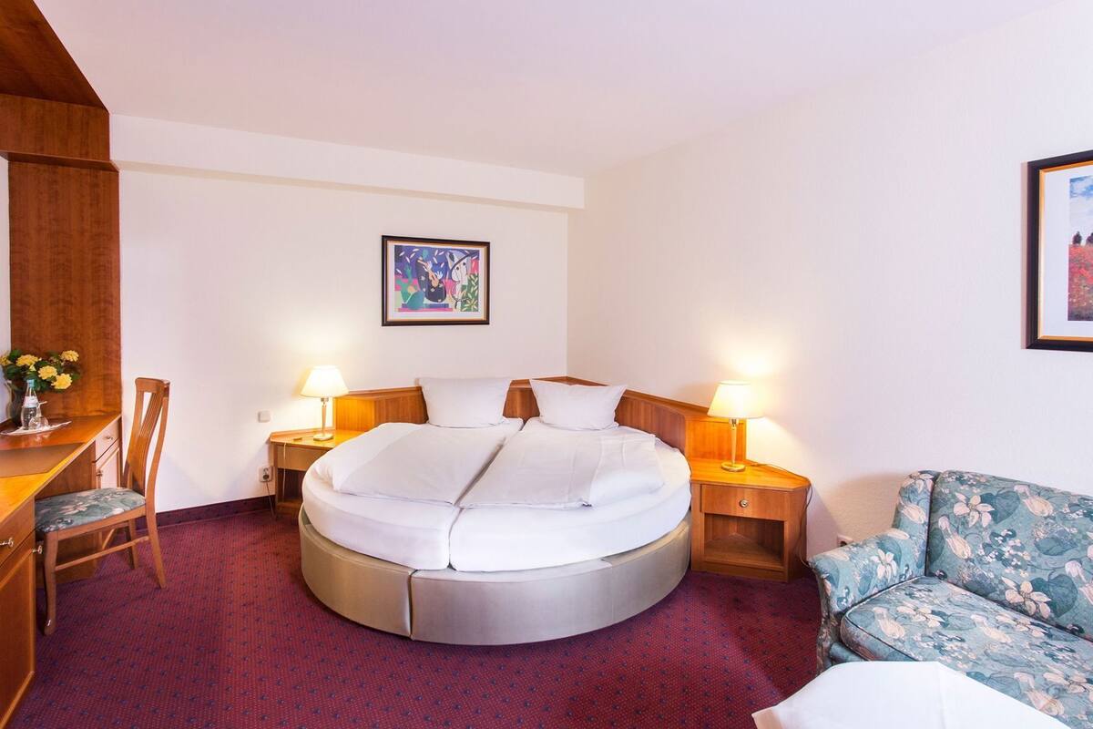 Quarto de hotel com uma cama redonda, duas mesas de cabeceira com luminárias, uma escrivaninha de madeira com uma cadeira e um sofá estampado com flores.