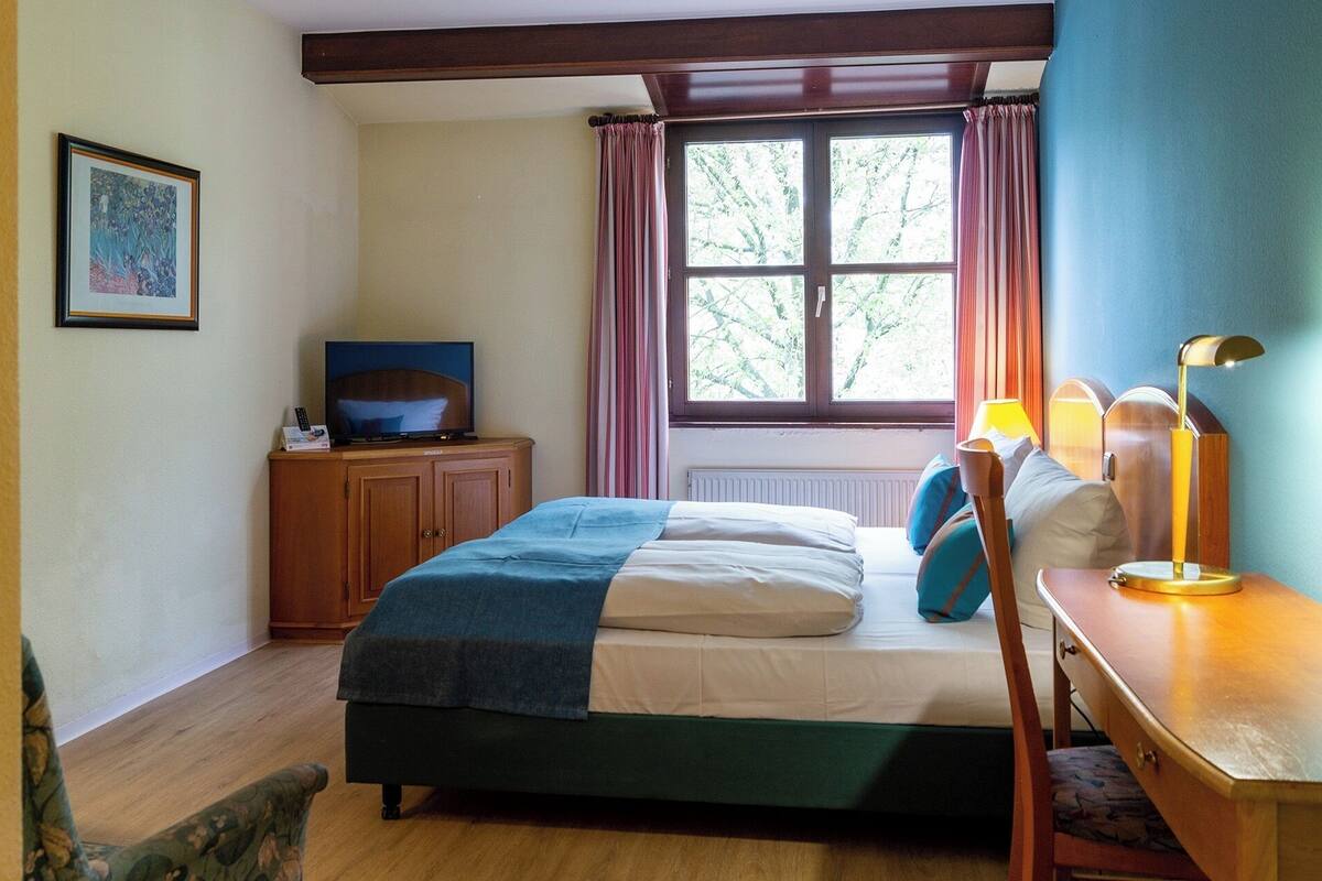Quarto com cama, televisão em aparador de madeira, janela com cortinas e escrivaninha com luminária.