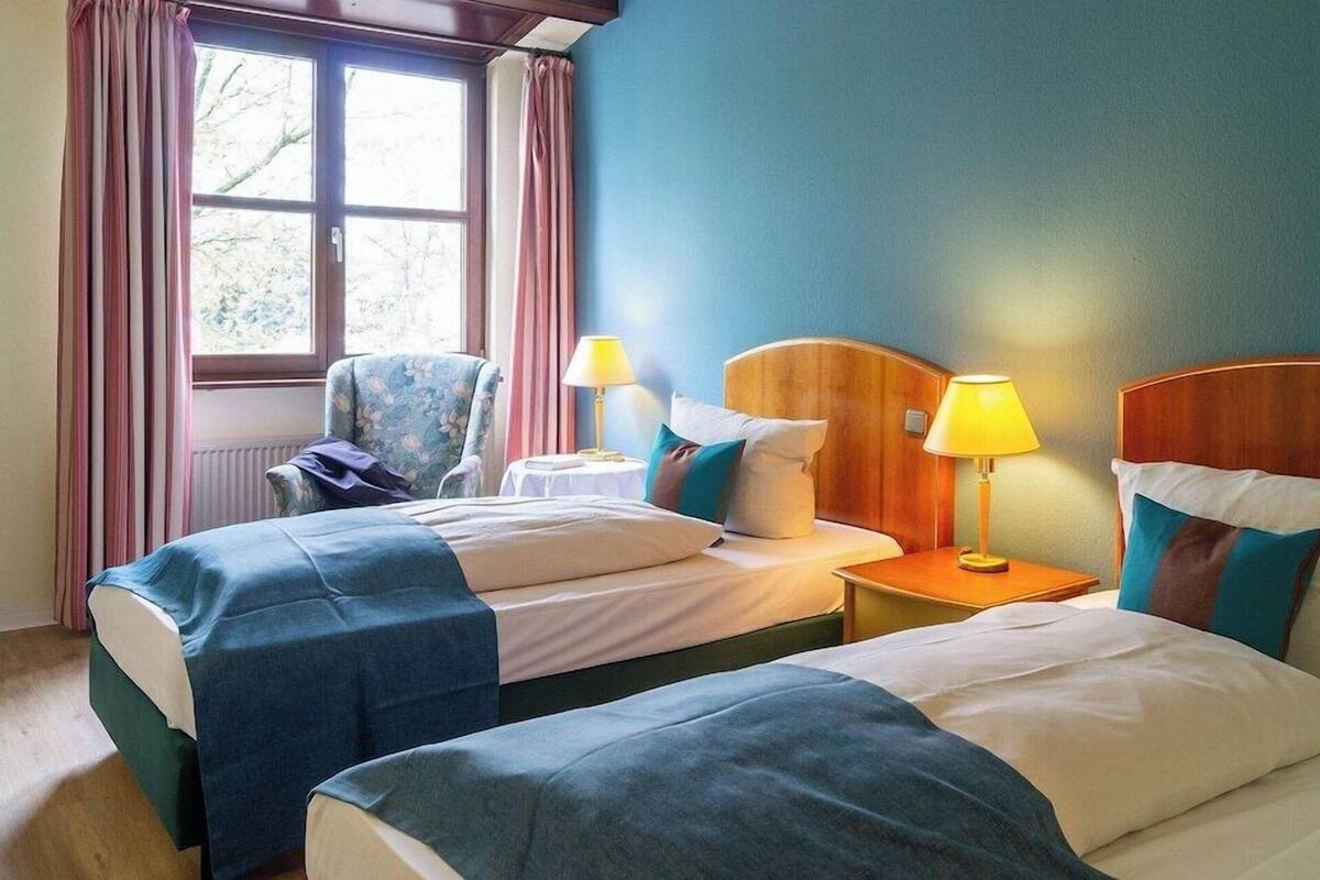 Quarto de hotel com duas camas, uma cadeira, uma janela com cortinas e duas luminárias de cabeceira.