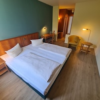 Cama bem arrumada com roupa de cama branca, cabeceira de madeira e uma pequena mesa de cabeceira com abajur.
