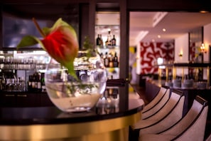 Uma área de bar com uma taça de vidro contendo uma flor e uma garrafa. Cadeiras e prateleiras com garrafas ao fundo.