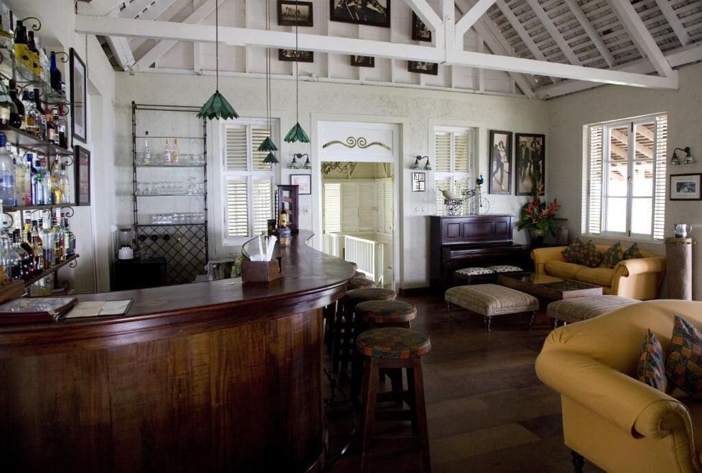 Uma área de bar com banquetas, um piano e um sofá numa sala com piso de madeira e paredes brancas.
