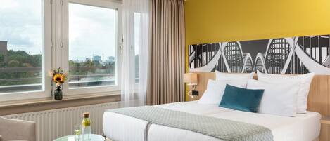 Quarto de hotel com uma cama grande, uma mesa de vidro com uma garrafa e um copo, uma cadeira, vista da cidade por grandes janelas e uma pintura de paisagem urbana na parede.