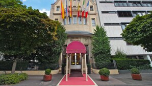 Entrada de hotel com tapete vermelho, várias bandeiras e estilo arquitetônico clássico.