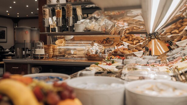 Buffet com diversos alimentos e recipientes, incluindo pão, doces e bebidas.