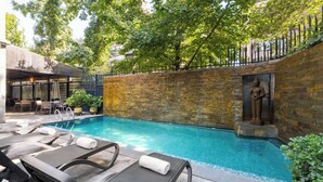 Uma piscina com espreguiçadeiras e uma estátua, cercada por um muro de pedra e vegetação.