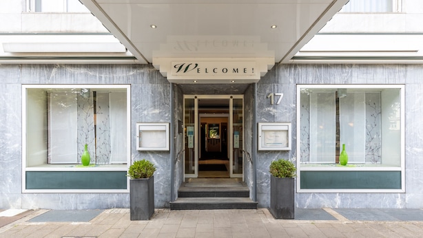 Uma entrada de hotel moderna com um letreiro 'Welcome!', janelas amplas e plantas em vasos.