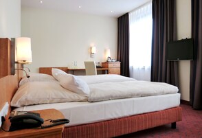 Quarto de hotel com cama, mesa de cabeceira com telefone, abajur e televisão na parede.