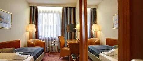 Quarto de hotel com duas camas, uma escrivaninha, uma cadeira, uma televisão e uma janela com cortinas.