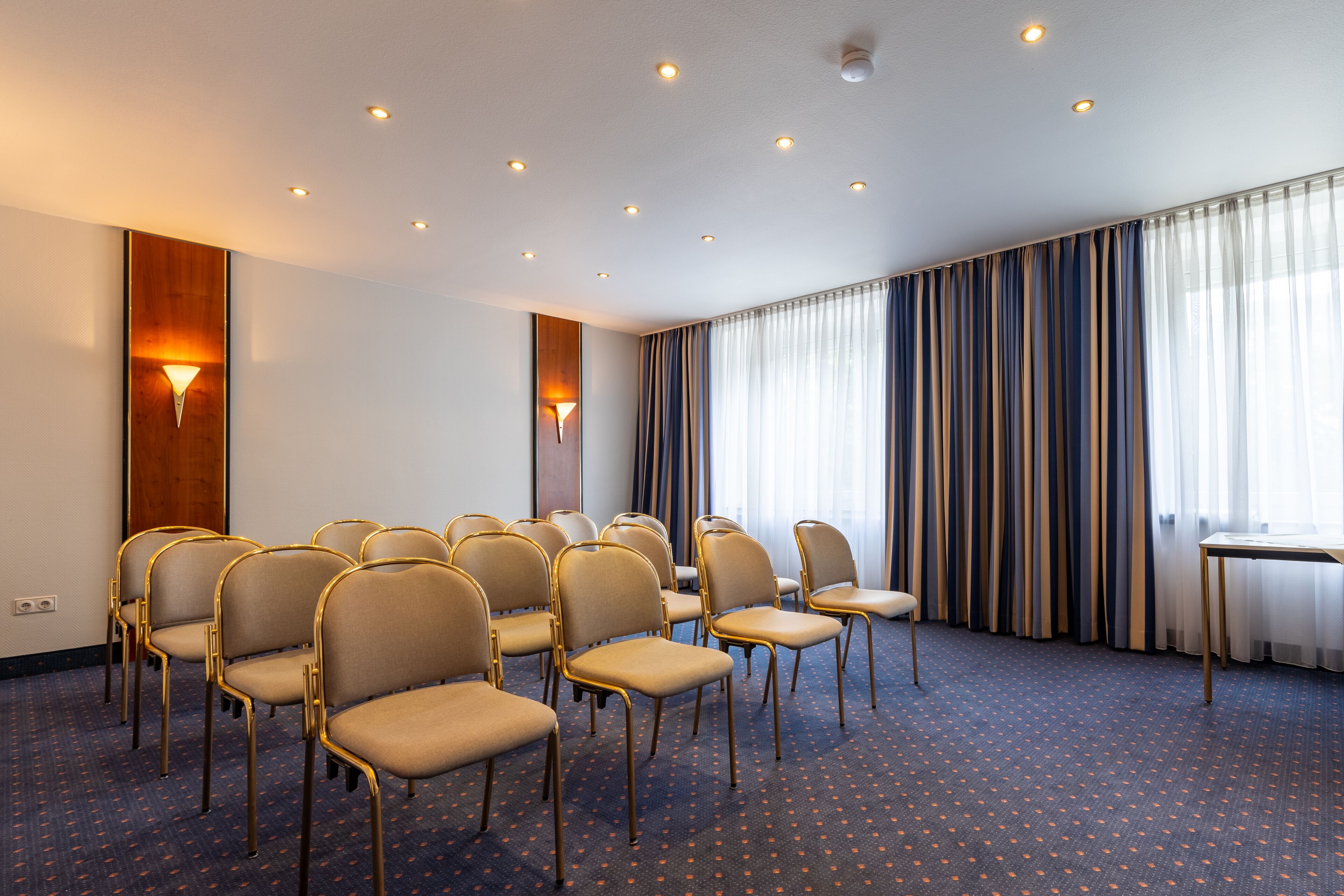 Sala de conferência com fileiras de cadeiras bege, uma parede de destaque em madeira e uma grande janela com cortinas.