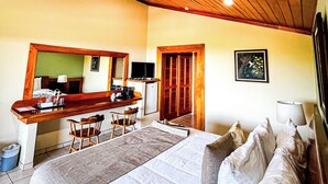 Quarto de hotel com teto de madeira, uma cama grande com roupa de cama branca, uma escrivaninha com computador, uma cadeira e uma pequena copa.