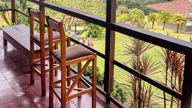 Um deck de madeira com um banco e cadeiras, com vista para uma paisagem exuberante e colinas distantes, e uma casa com telhado vermelho.