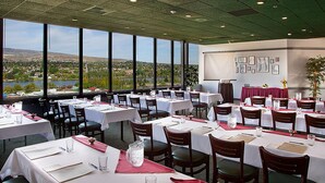 Restaurante com mesas preparadas para refeições, grandes janelas com vista para um lago e uma cidade, e quadros nas paredes.