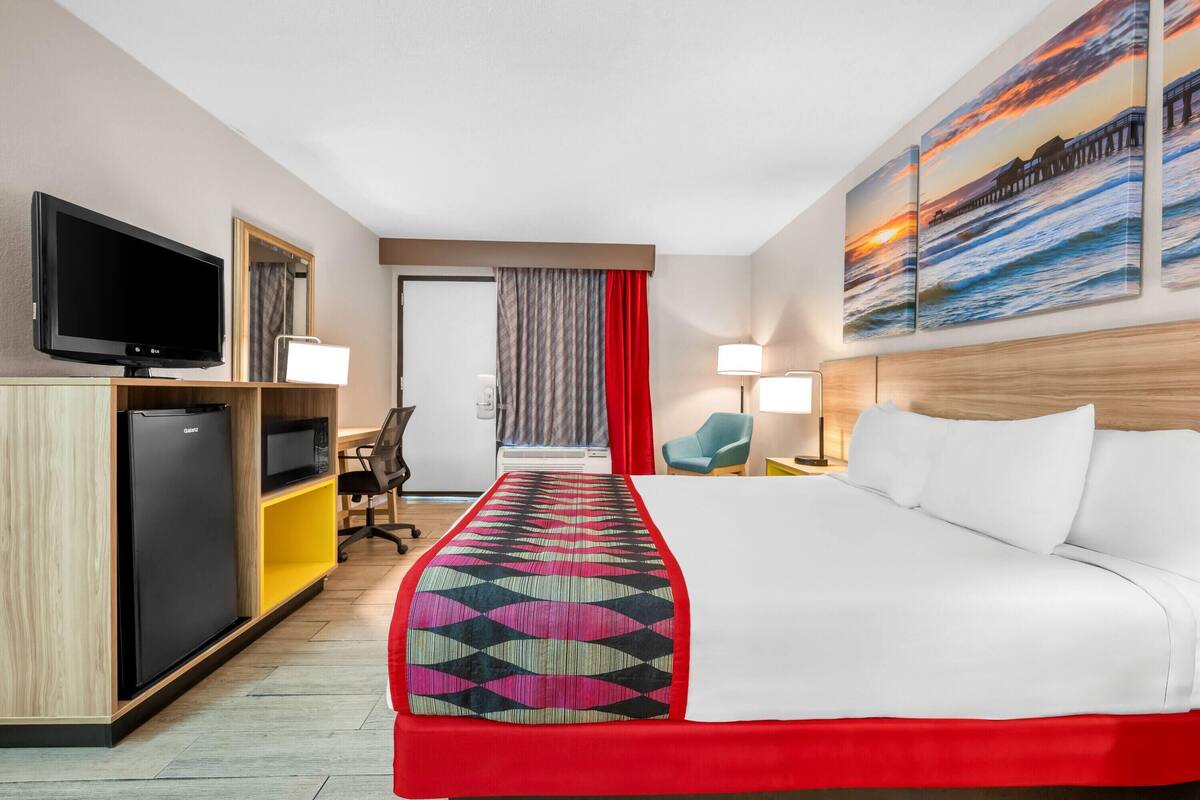 Quarto de hotel com cama, televisão, frigorífico, secretária com cadeira e pintura de um pôr do sol sobre o oceano.