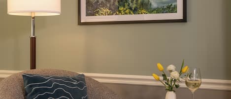 Quarto com pintura emoldurada de uma paisagem, luminária, cadeira com almofada estampada, mesa pequena com um vaso de flores e uma taça de vinho, e um livro.