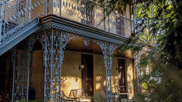 Varanda com grades de metal ornamentadas, porta de madeira e área de estar com cadeiras e mesa.