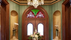 Um amplo corredor com piso quadriculado, portas de madeira ornamentadas e um candelabro.