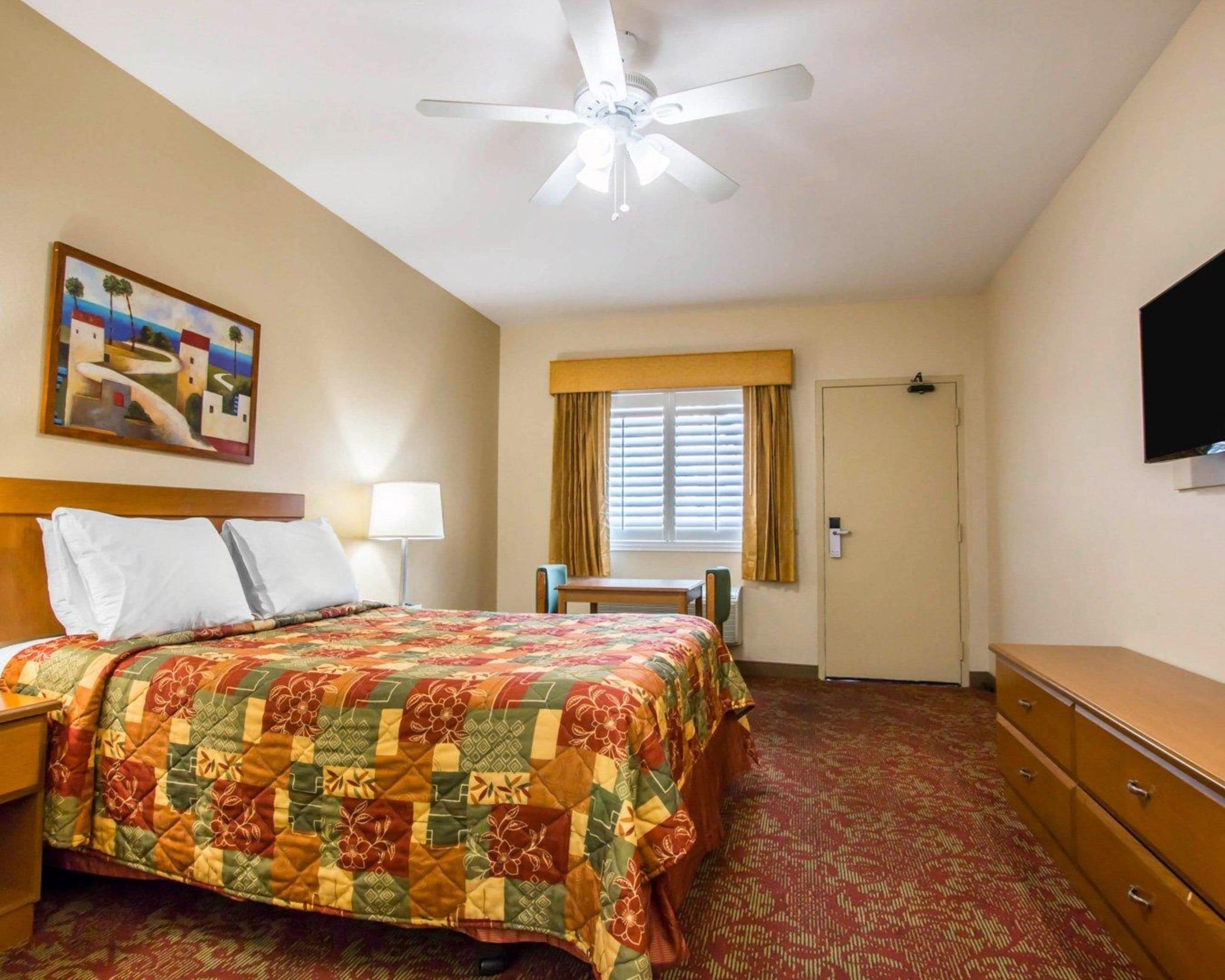 Quarto de hotel com cama, escrivaninha, cadeira, ventoinha no teto, um quadro na parede e televisão.