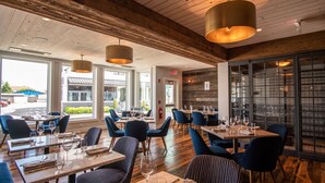 Restaurante com vigas de madeira, grandes janelas e cadeiras azuis. Mesas montadas com copos de vinho e talheres.