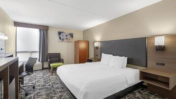 Quarto de hotel com uma cama grande, uma mesa de trabalho com cadeira, um guarda-roupa e um quadro na parede.