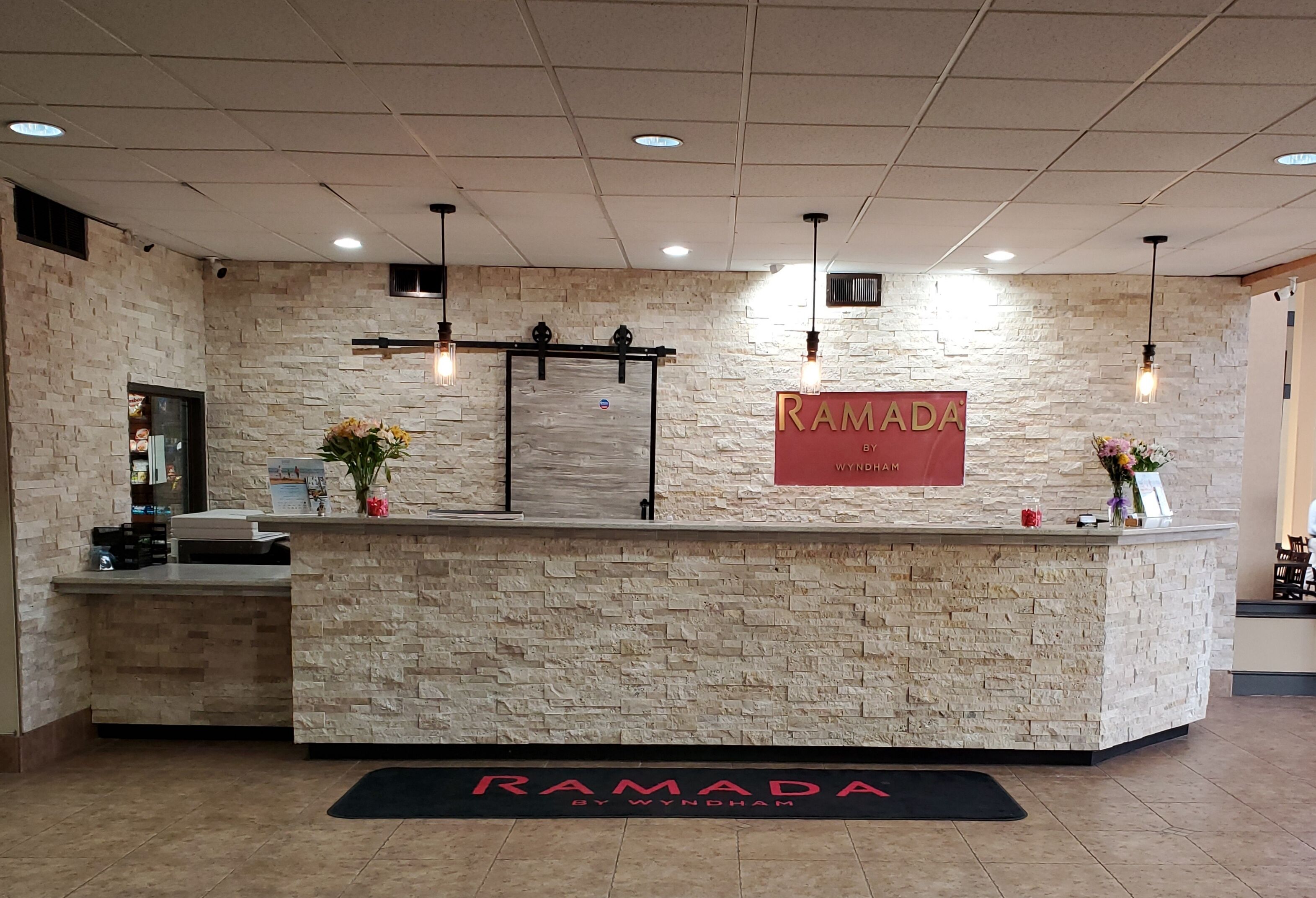 Uma área de recepção com parede de pedra, uma placa indicando "RAMADA BY WYNDHAM" e um tapete preto no chão.