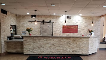 Uma área de recepção com parede de pedra, uma placa indicando "RAMADA BY WYNDHAM" e um tapete preto no chão.