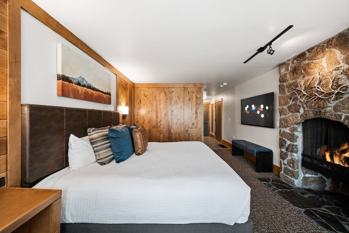 Quarto com uma cama grande, cabeceira de madeira, lareira de pedra e um quadro na parede.