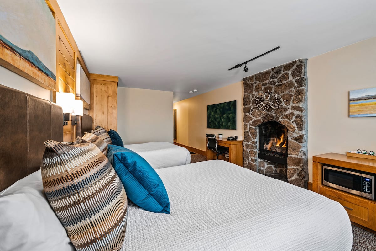 Quarto de hotel com lareira de pedra, cama com roupa de cama branca, escrivaninha com computador e cabeceira de madeira.