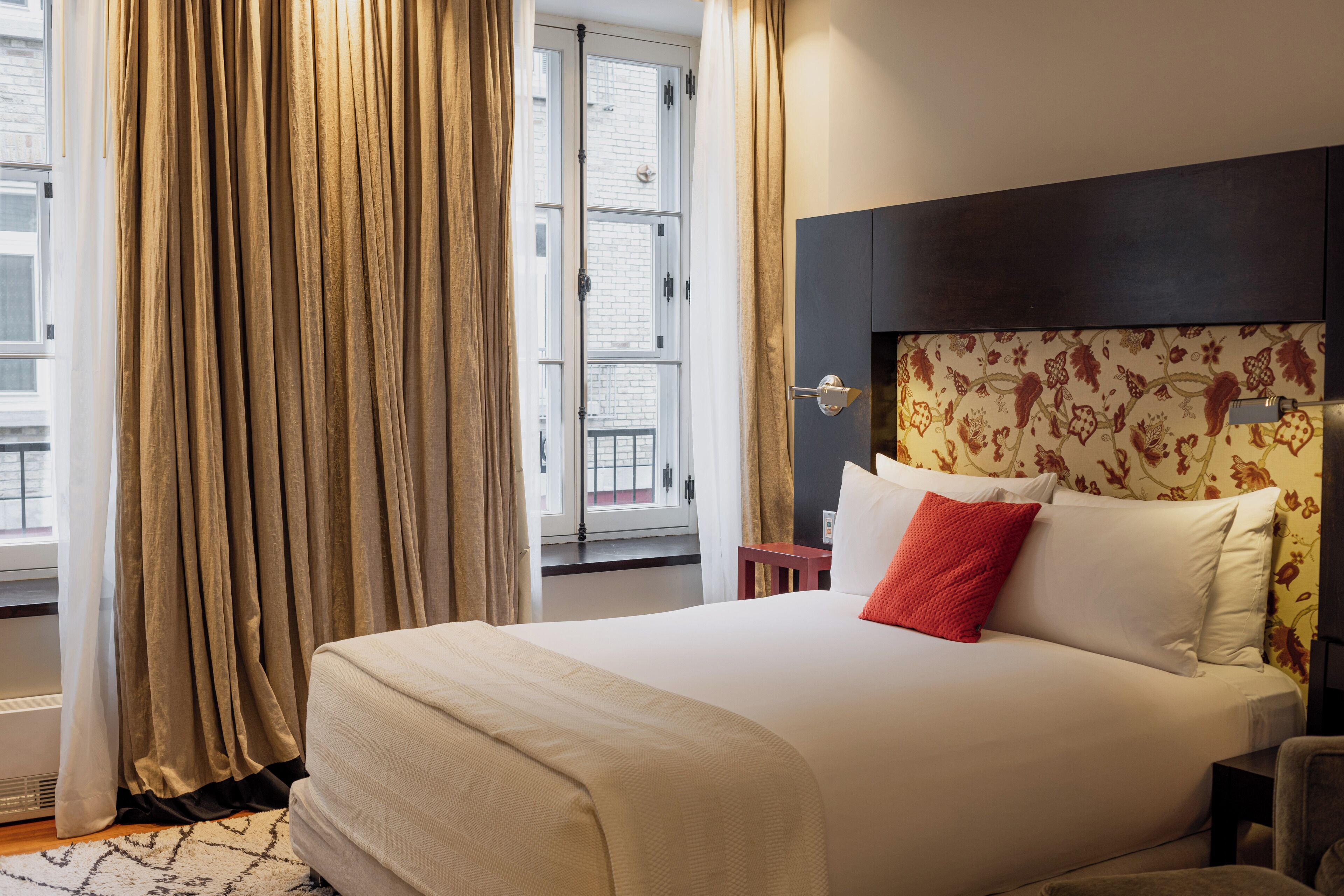 Quarto de hotel com uma cama grande, cabeceira com papel de parede floral, janela com cortinas e vista para edifícios.