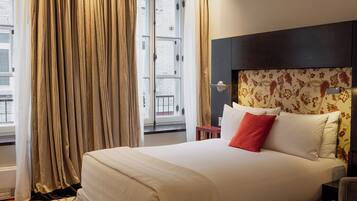 Quarto de hotel com uma cama grande, cabeceira com papel de parede floral, janela com cortinas e vista para edifícios.