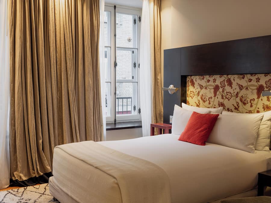 Quarto de hotel com uma cama grande, cabeceira com papel de parede floral, janela com cortinas e vista para edifícios.