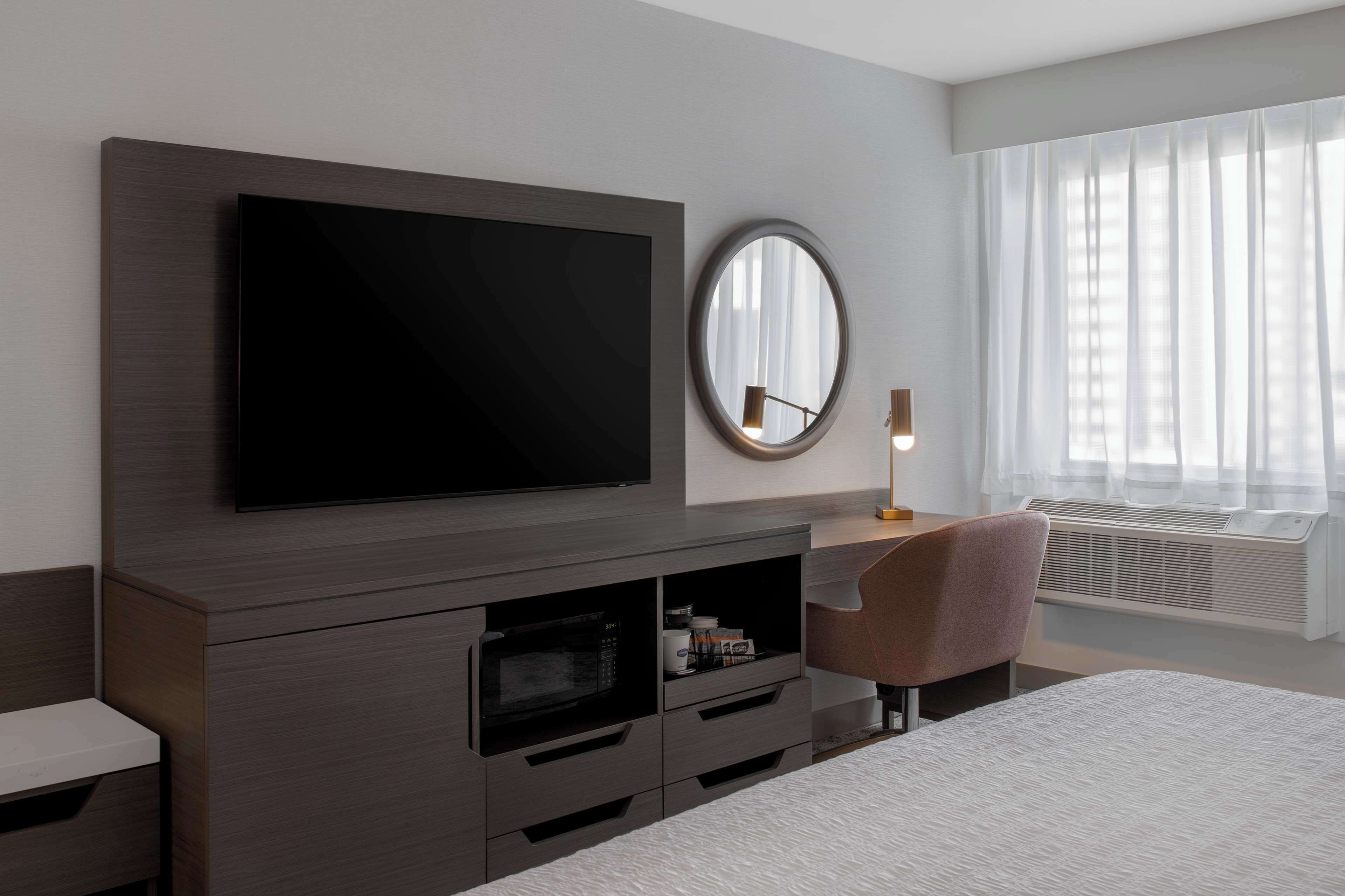 Quarto de hotel moderno com televisão de tela plana fixada em um armário de madeira, um espelho redondo, mesa de cabeceira, uma cadeira e uma janela grande com cortinas translúcidas.