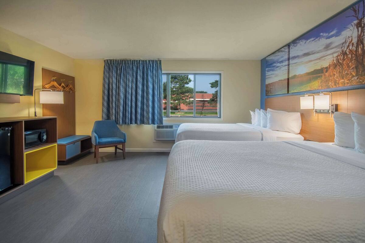Quarto de hotel com duas camas, uma escrivaninha com televisão, uma cadeira, uma janela com cortinas e um quadro na parede.